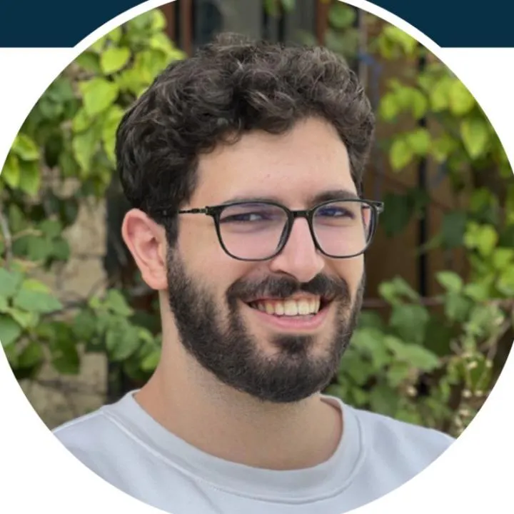 Man with dark curly hair, beard, and glasses smiling in front of green ...